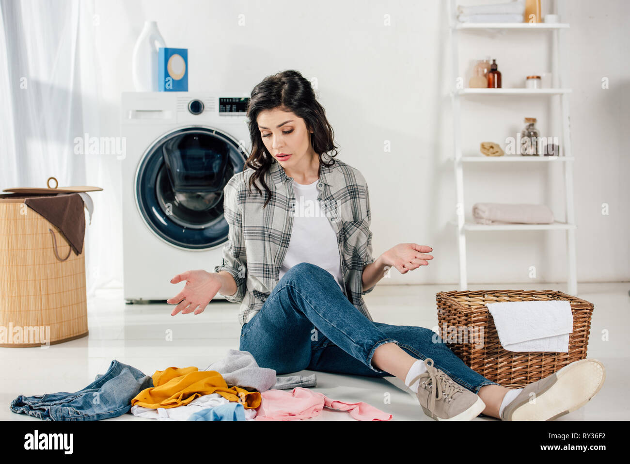 confused woman sitting on floor near scattered clothes and baskets in ...