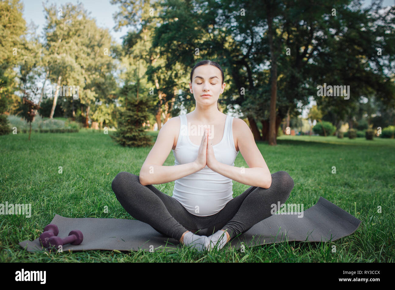 Young pregnant woman sit on yoga mate and pray outside in park. She ...