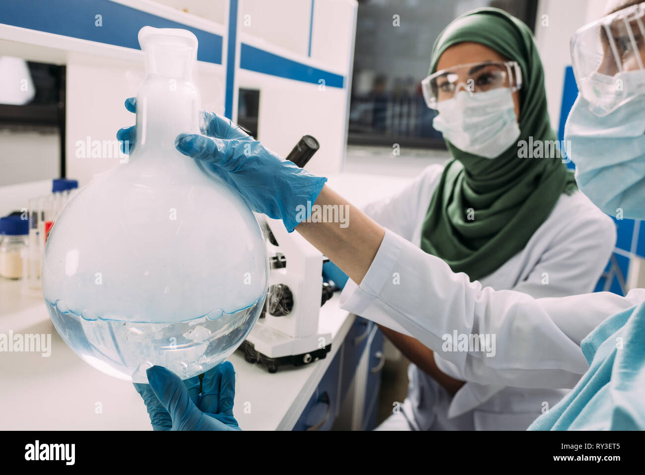 female muslim scientists in goggles holding flask with liquid while ...