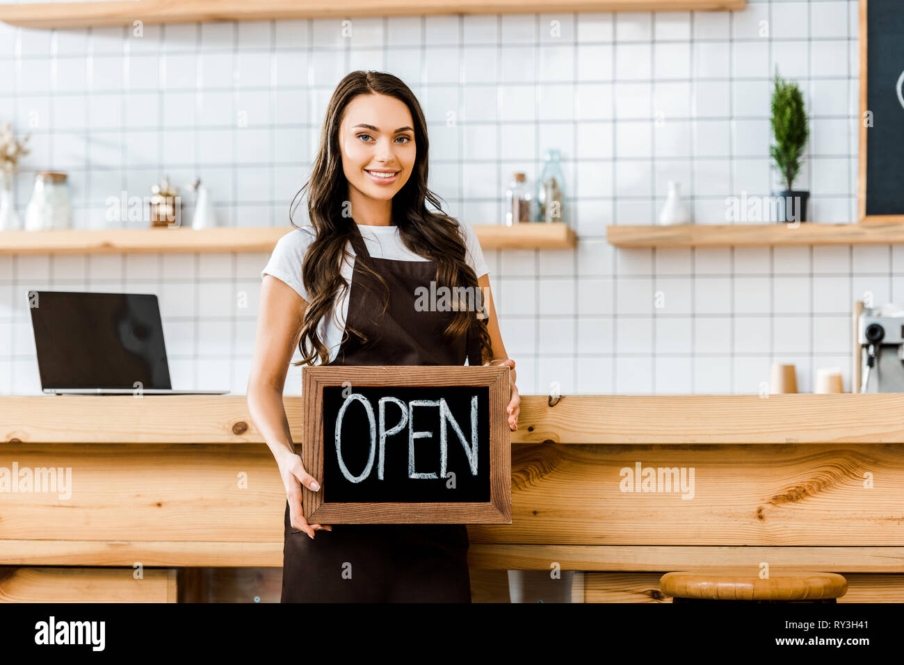 attractive cashier in brown apron standing near wooden bar counter and ...