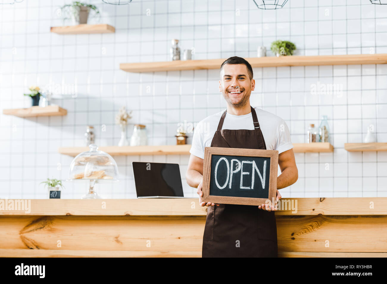 handsome cashier in brown apron standing near wooden bar counter and ...