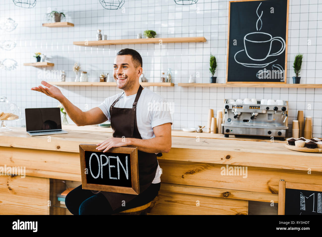 handsome cashier in apron sitting near wooden bar counter, holding ...