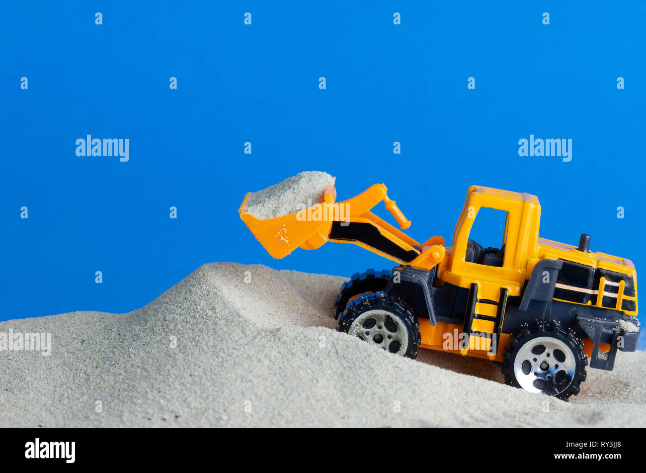 Yellow toy tractor is digging and lift sand on the blue background ...