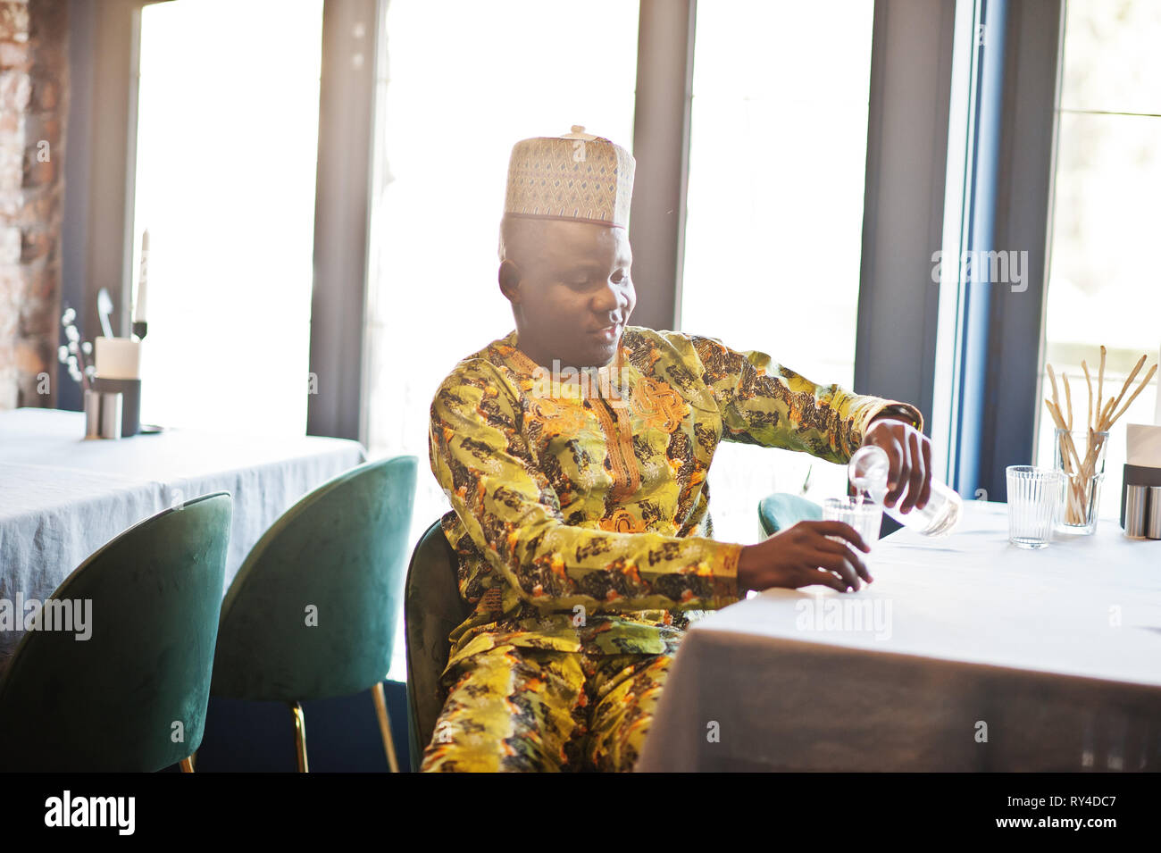 Friendly afro man in traditional clothes with cap sitting at restaurant ...