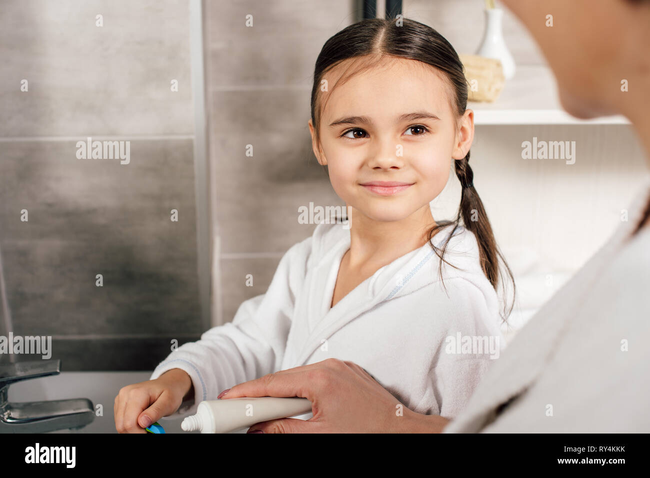 mother applying toothpaste from tube on daughter toothbrush in bathroom ...