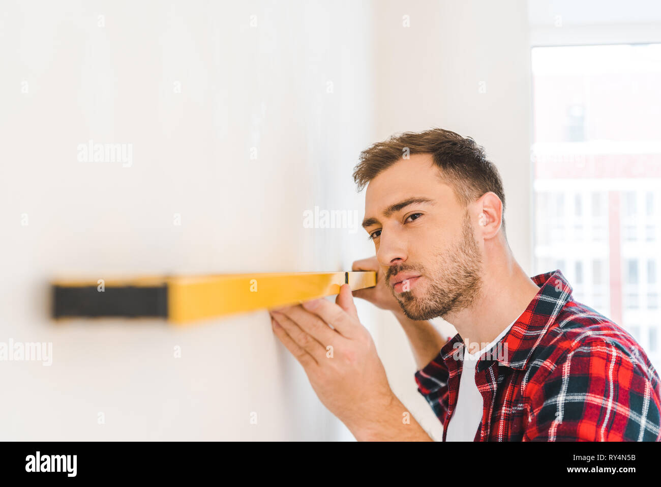handsome handyman holding measuring level while measuring wall at home ...