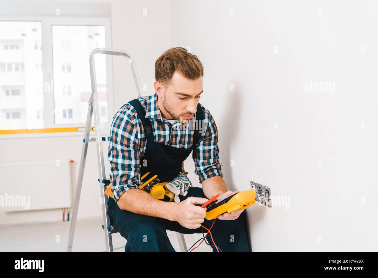 handsome handyman holding digital multimeter while sitting near power ...