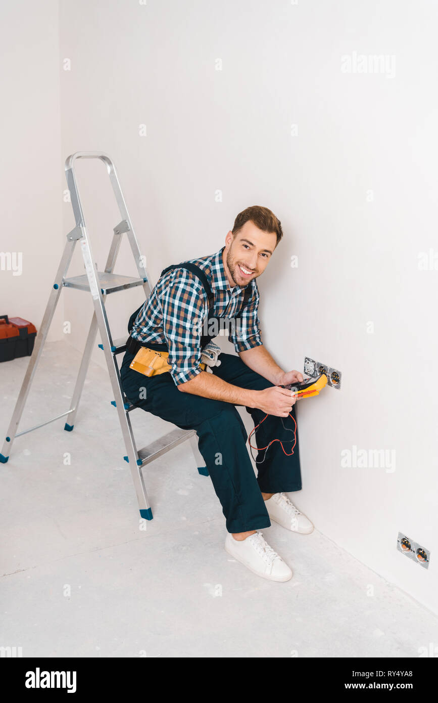 happy handyman holding digital multimeter and sitting on ladder near ...