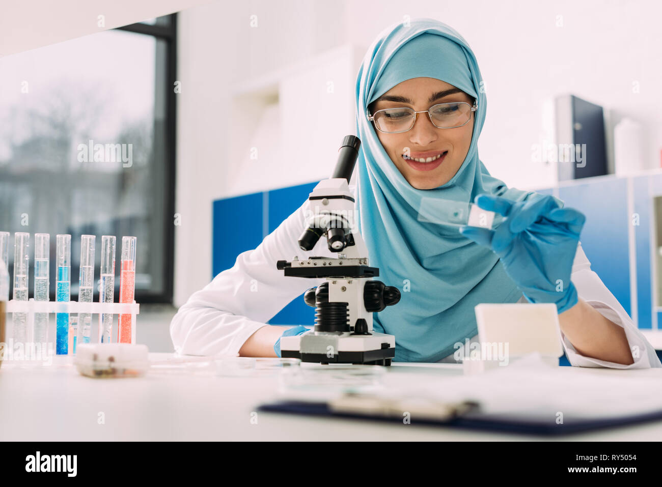 smiling female muslim scientist sitting at table with microscope and looking at glass sample ...