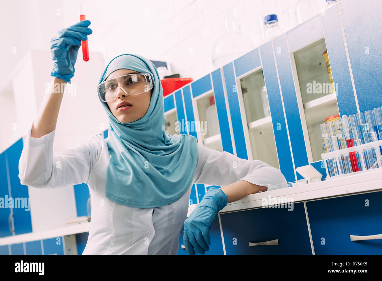 female muslim scientist in goggles holding test tube with red liquid in ...