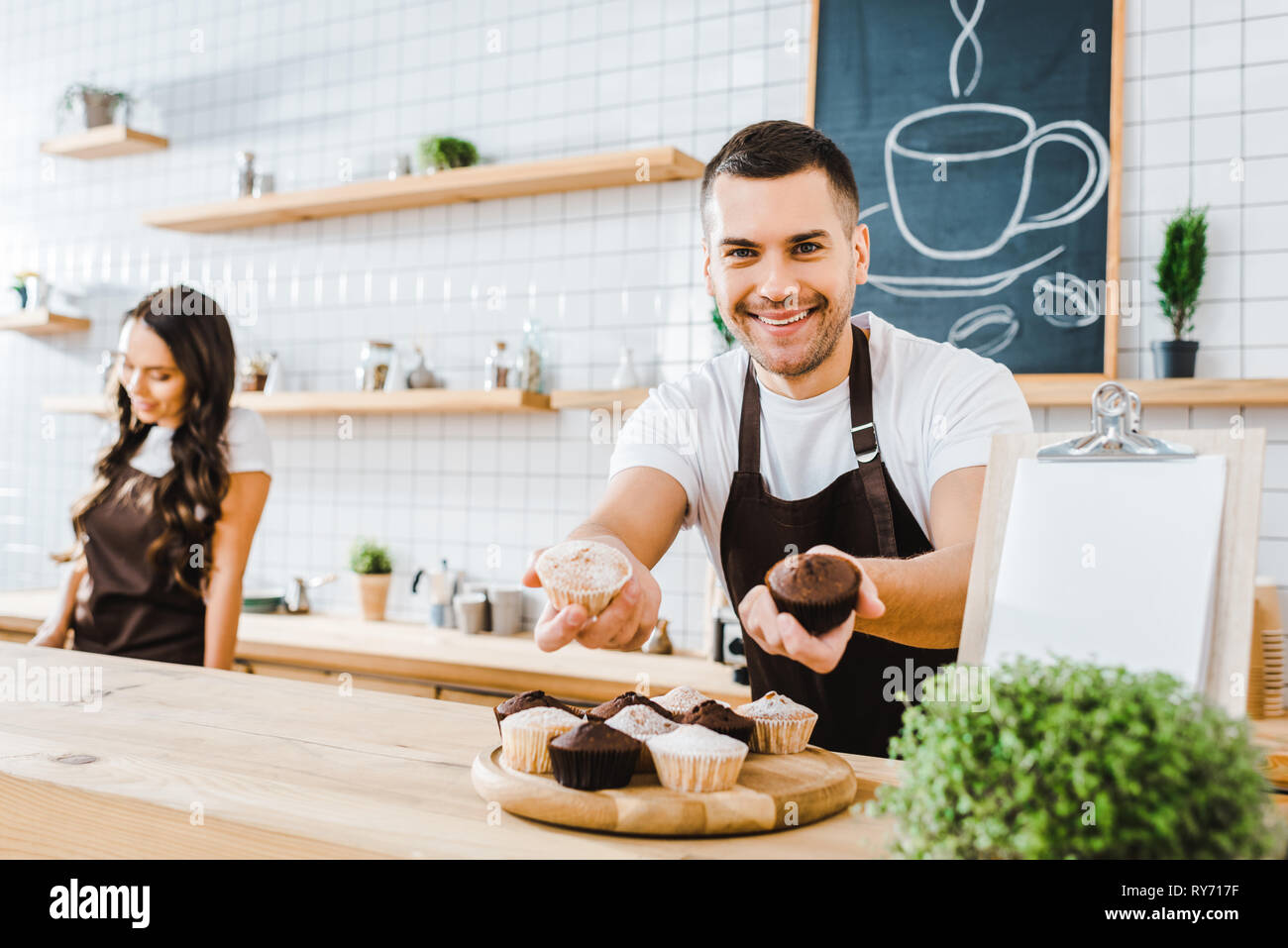 selective focus of handsome cashier in apron giving cupcakes wile ...
