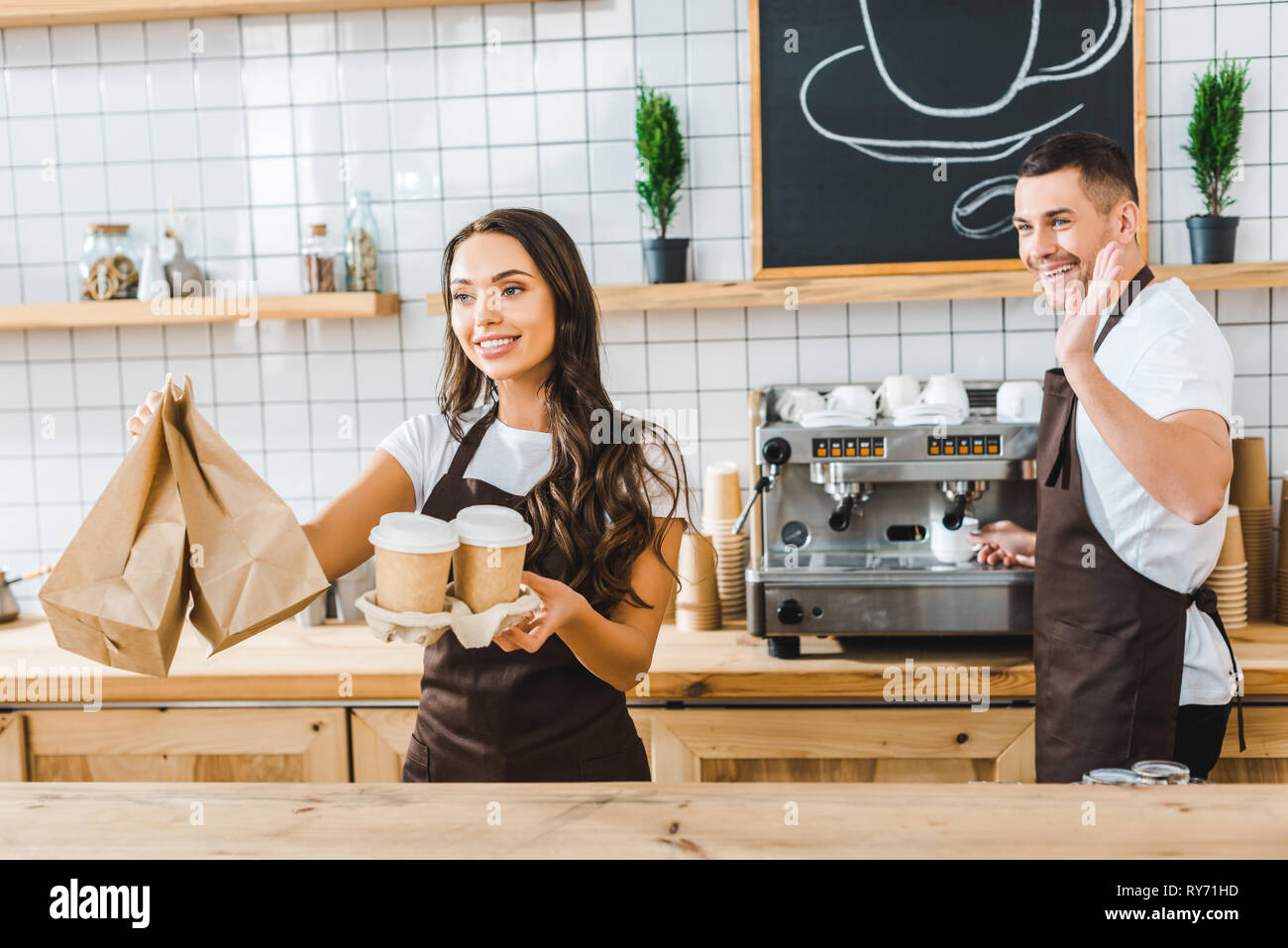 attractive brunette cashier holding paper cups and bags wile barista ...