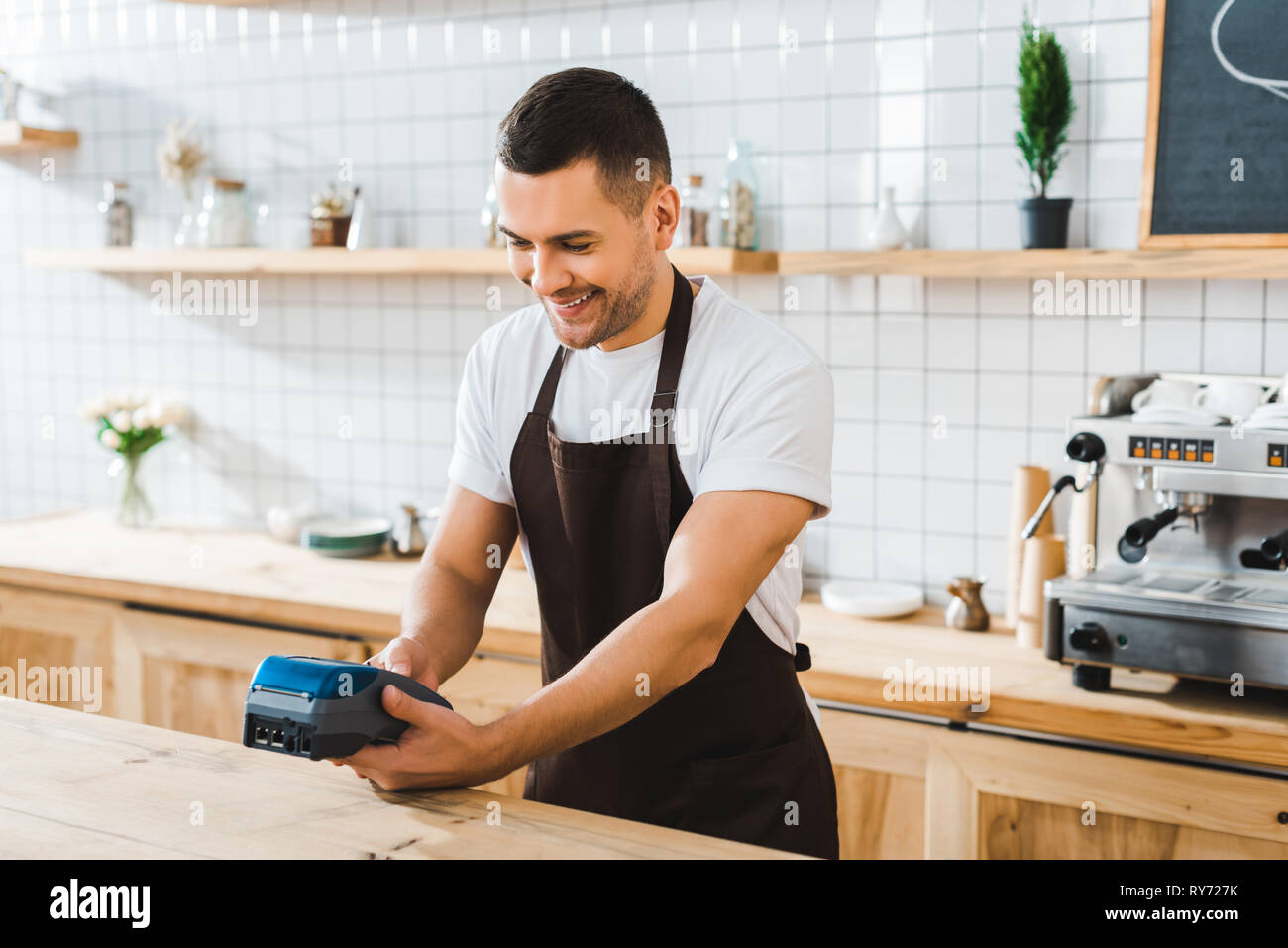 handsome cashier standing near bar counter in brown apron and holding ...