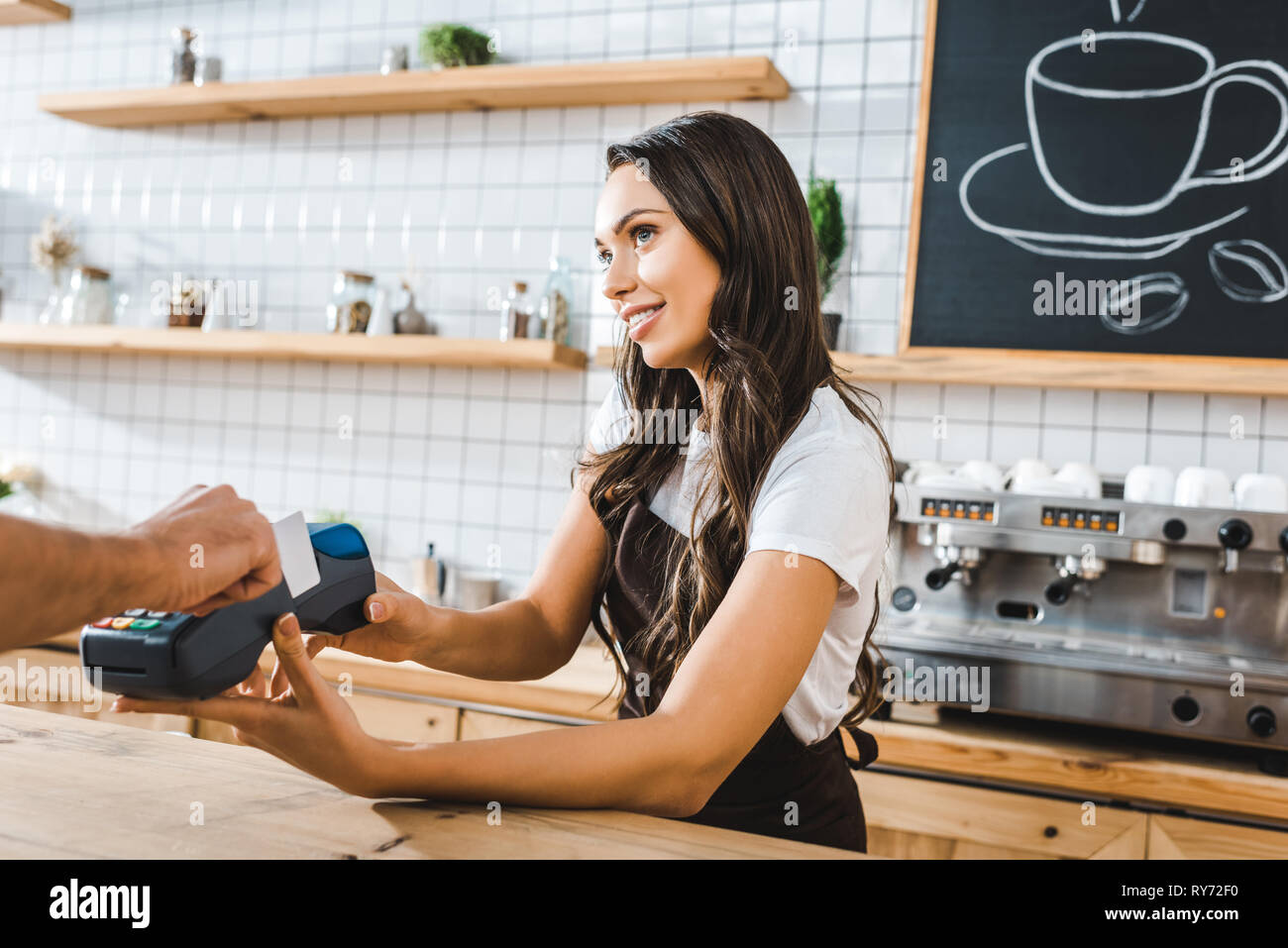attractive cashier standing near bar counter in brown apron and holding ...