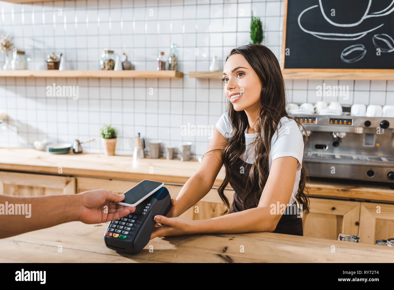 attractive cashier standing near bar counter in brown apron and holding ...