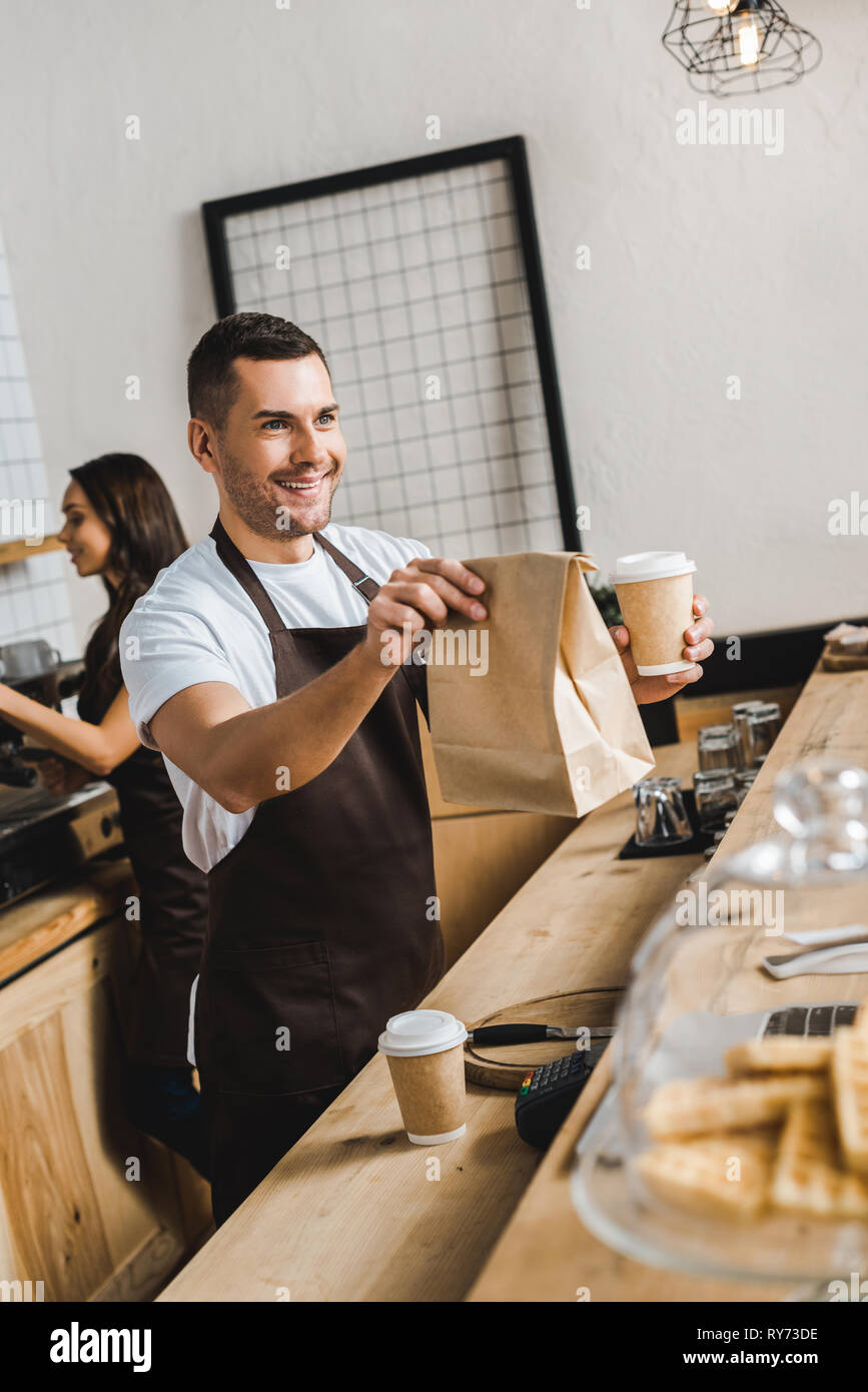 handsome cashier in brown apron giving paper cup and bag wile barista ...