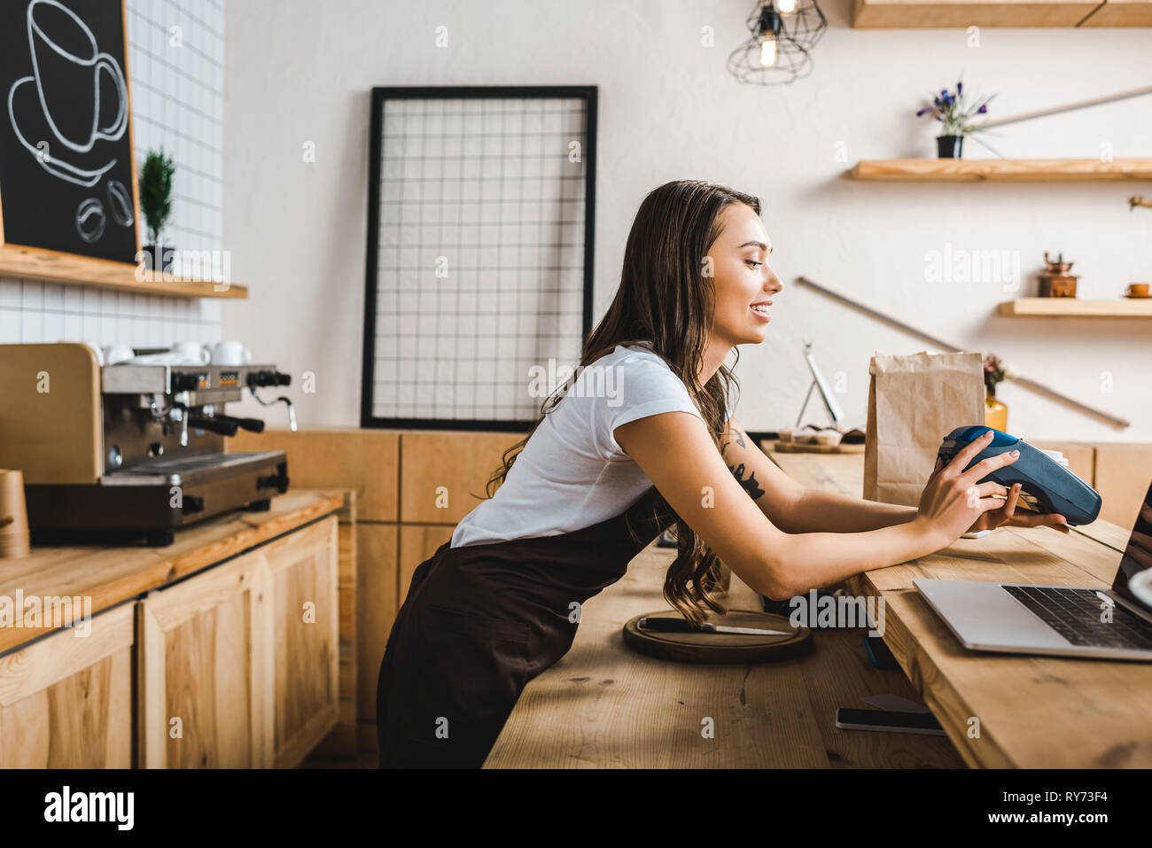 attractive cashier standing in brown apron and holding terminal behind ...