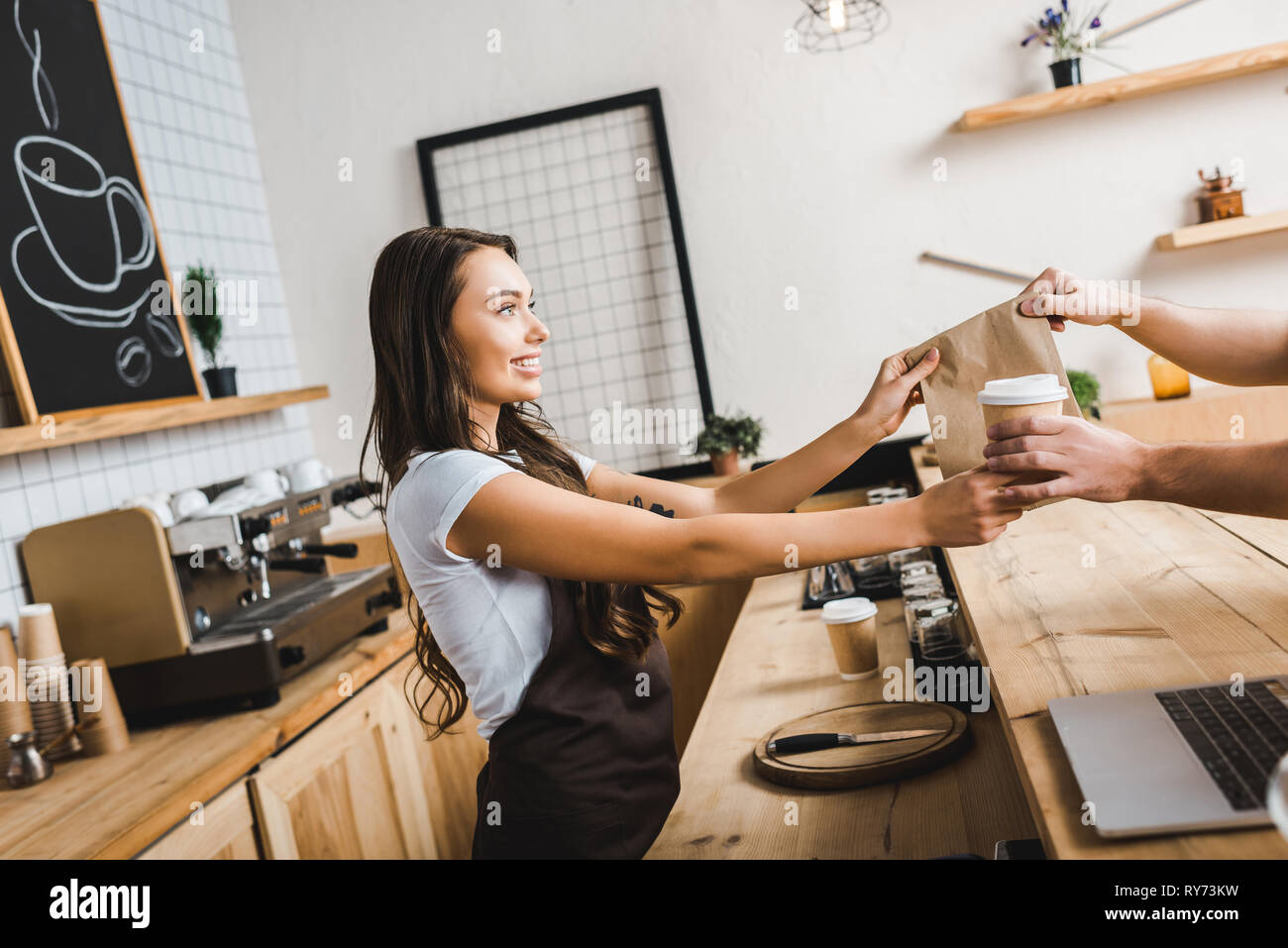 attractive cashier in brown apron giving paper cup and bag to man in ...