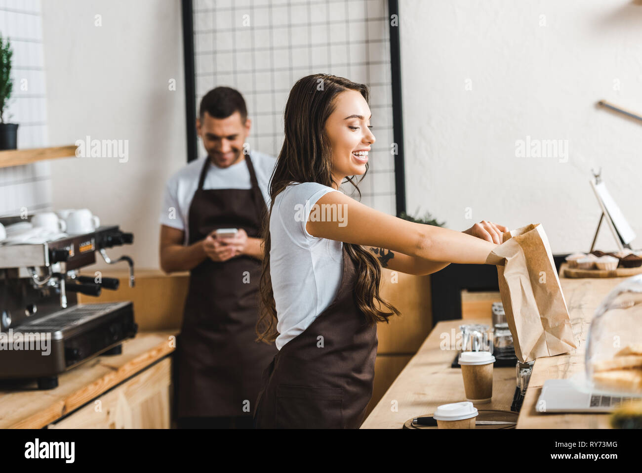 attractive cashier in brown apron packing paper bag wile barista ...