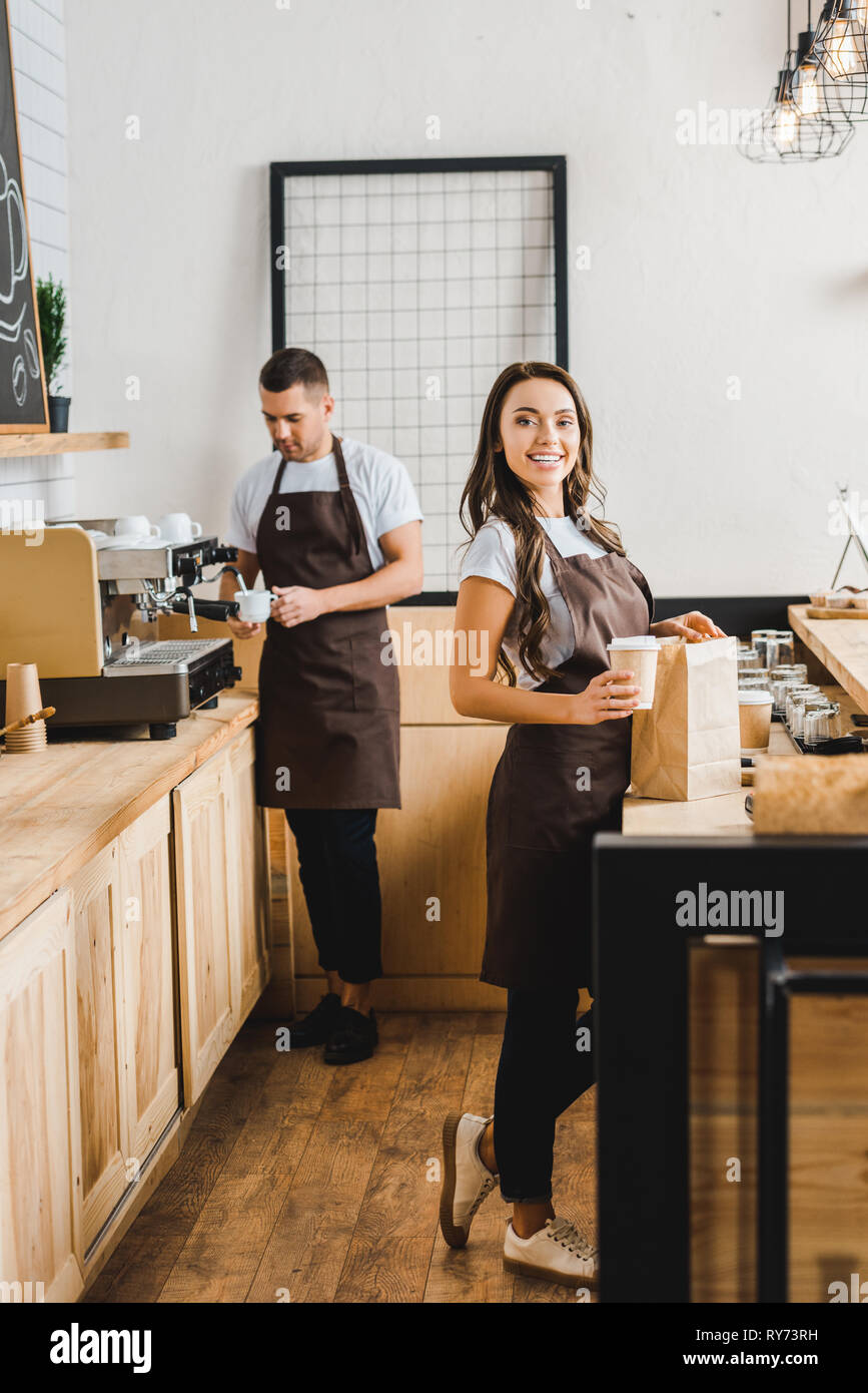 attractive cashier with paper cup and bag with barista in brown apron ...
