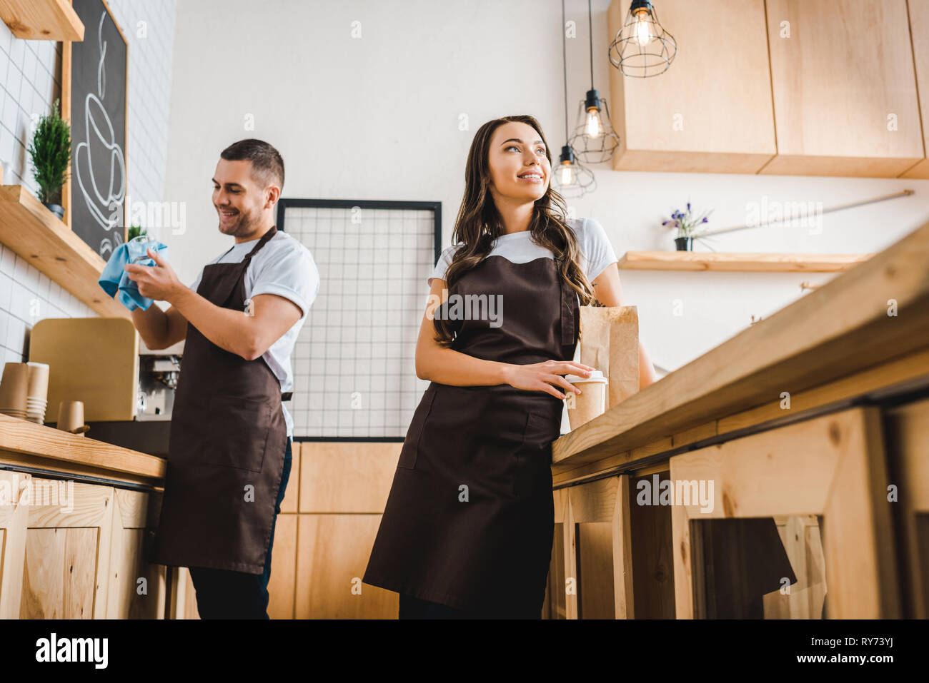barista and cashier in aprons standing behind bar counter in coffee ...