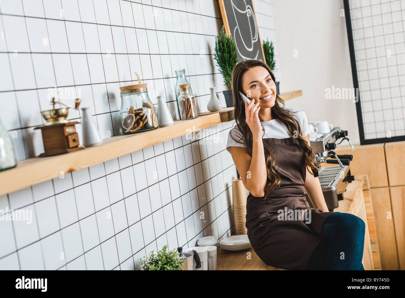 attractive cashier in brown apron talking on smartphone near coffee ...