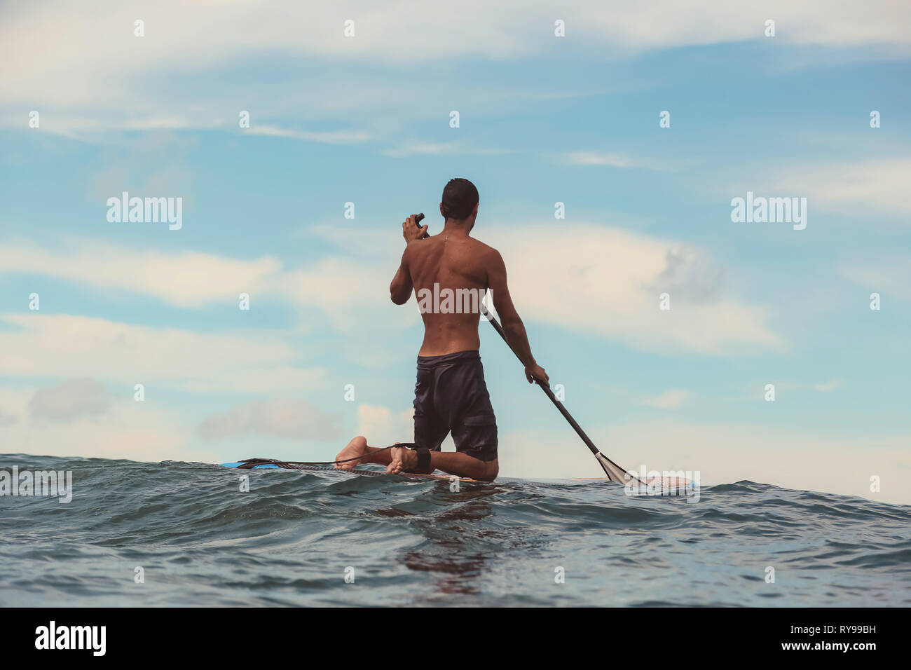 Back view of male paddling on surf board between water of sea and blue ...