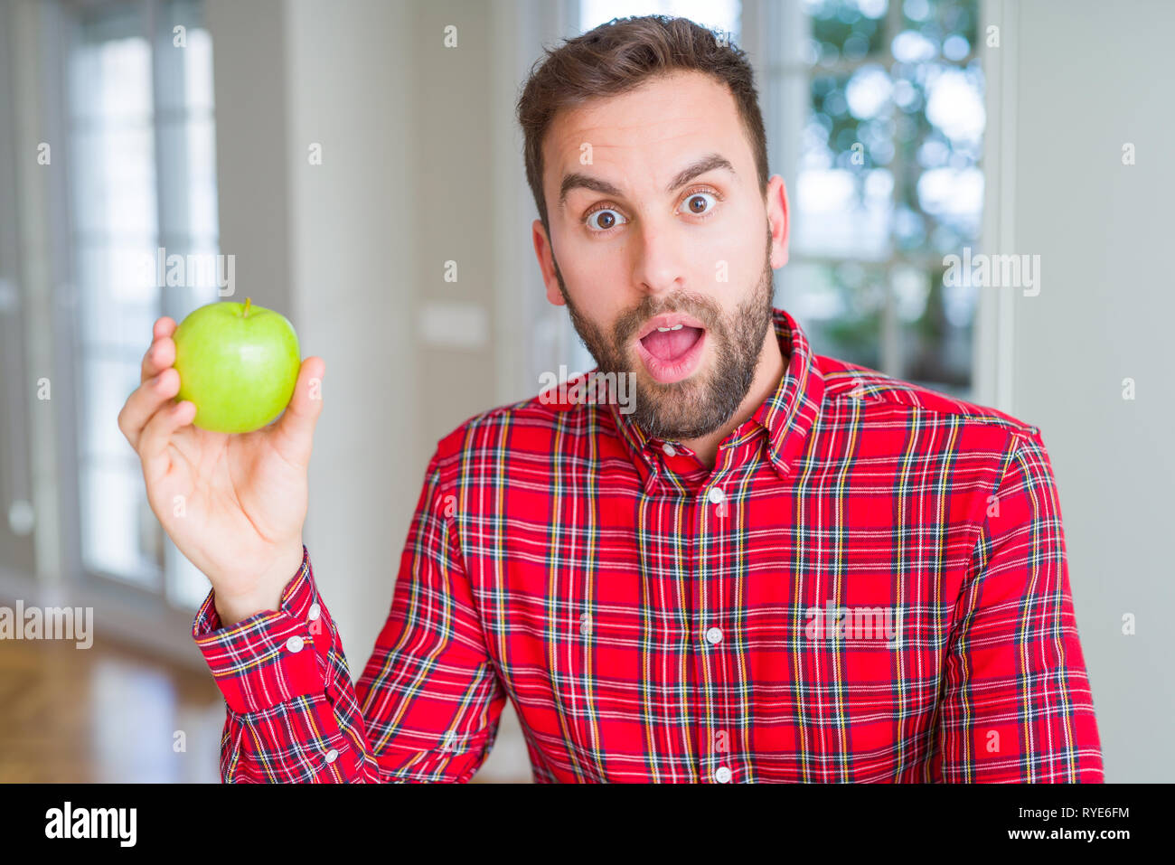 Handsome man eating fresh healthy green apple scared in shock with a ...