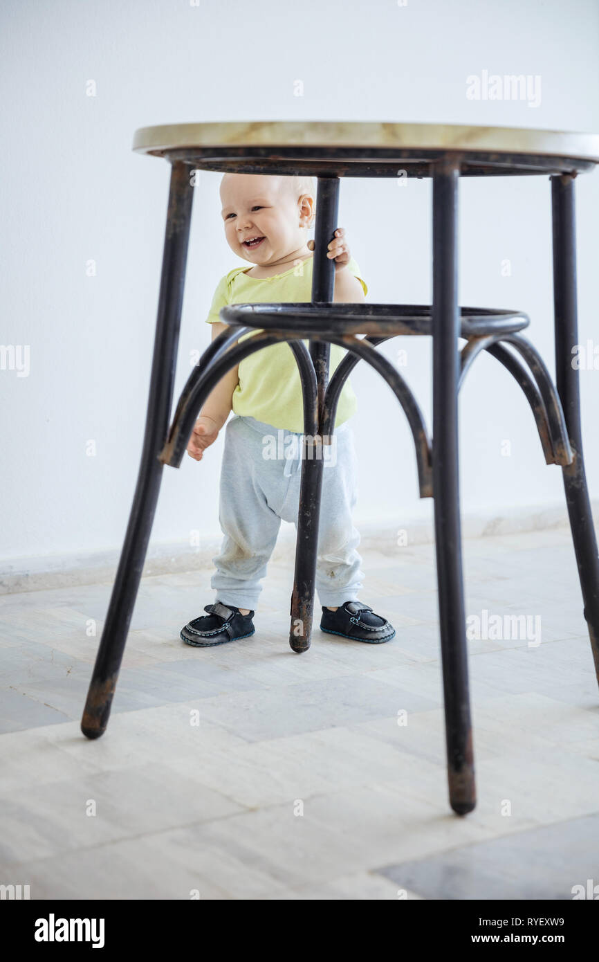 Cute baby girl standing while gripping leg of stool and laughing. Child ...