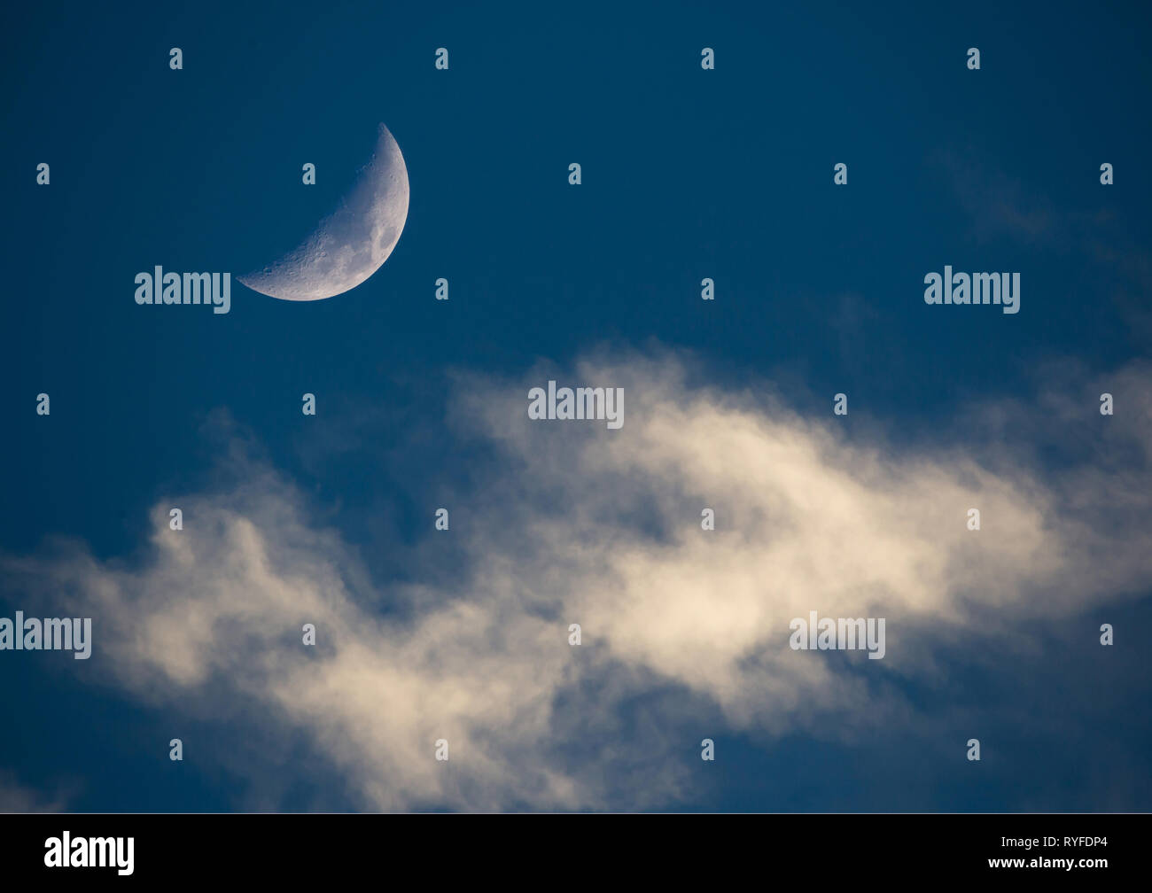 A setting sun illuminates a crescent moon and wispy clouds on a dark ...