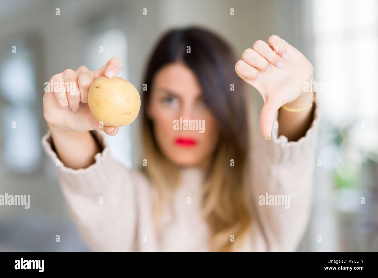Young beautiful woman holding fresh potato at home with angry face ...