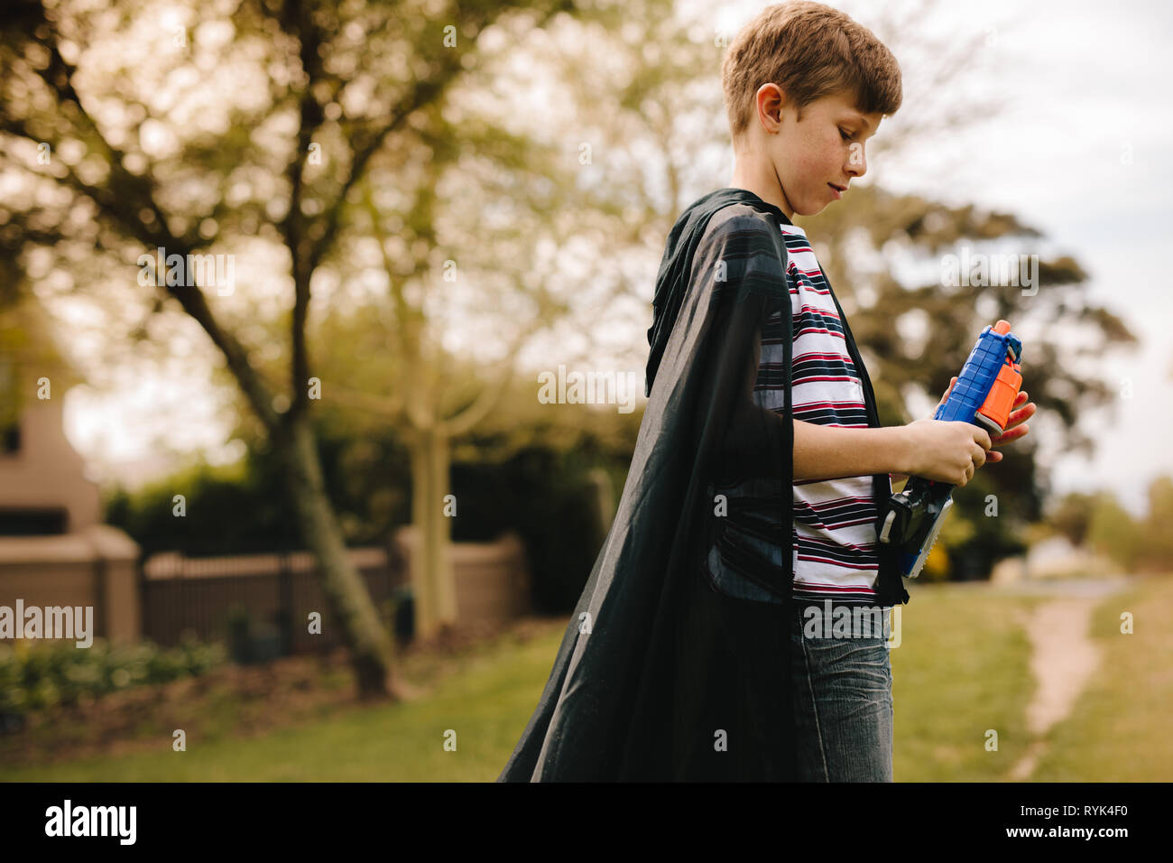 Side view of a young boys wearing a cape playing with toy gun outdoors ...
