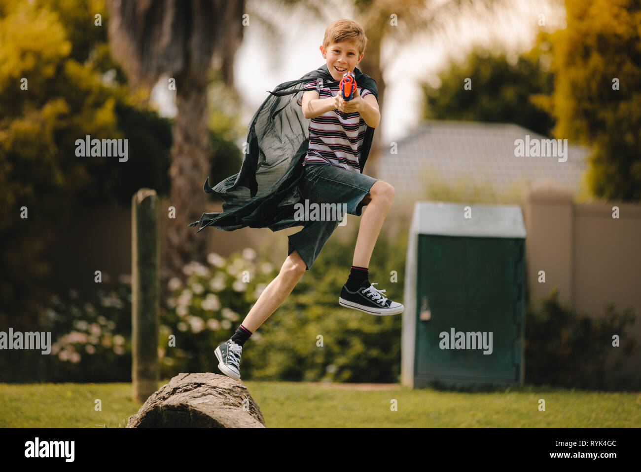 Young boy wearing cape jumping over a log with a toy gun. Boy ...