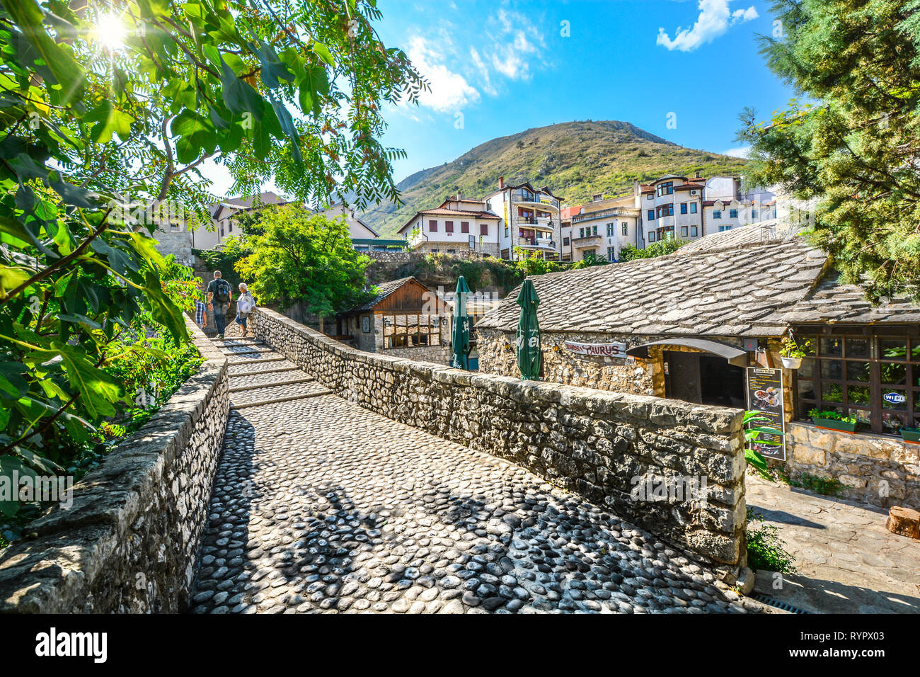Tourists walk a narrow cobblestone path over a bridge and past a cafe ...