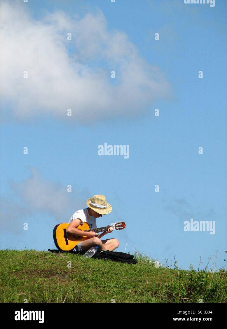 A seated guitarist in a straw hat playing his guitar on a grassy hill ...