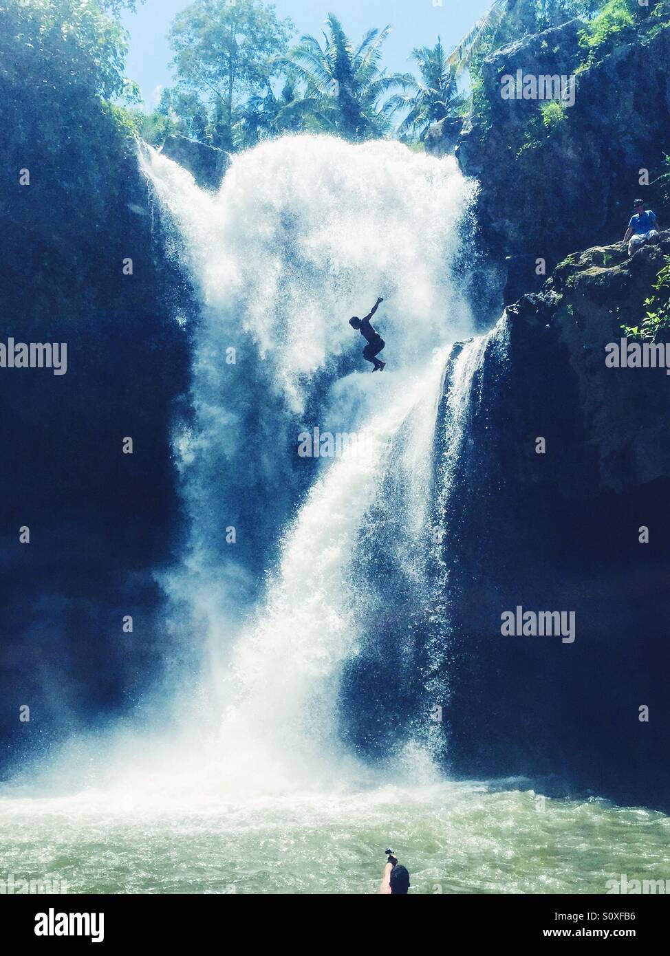 Man jumping from the cliff into the waterfall, while friend filming him ...