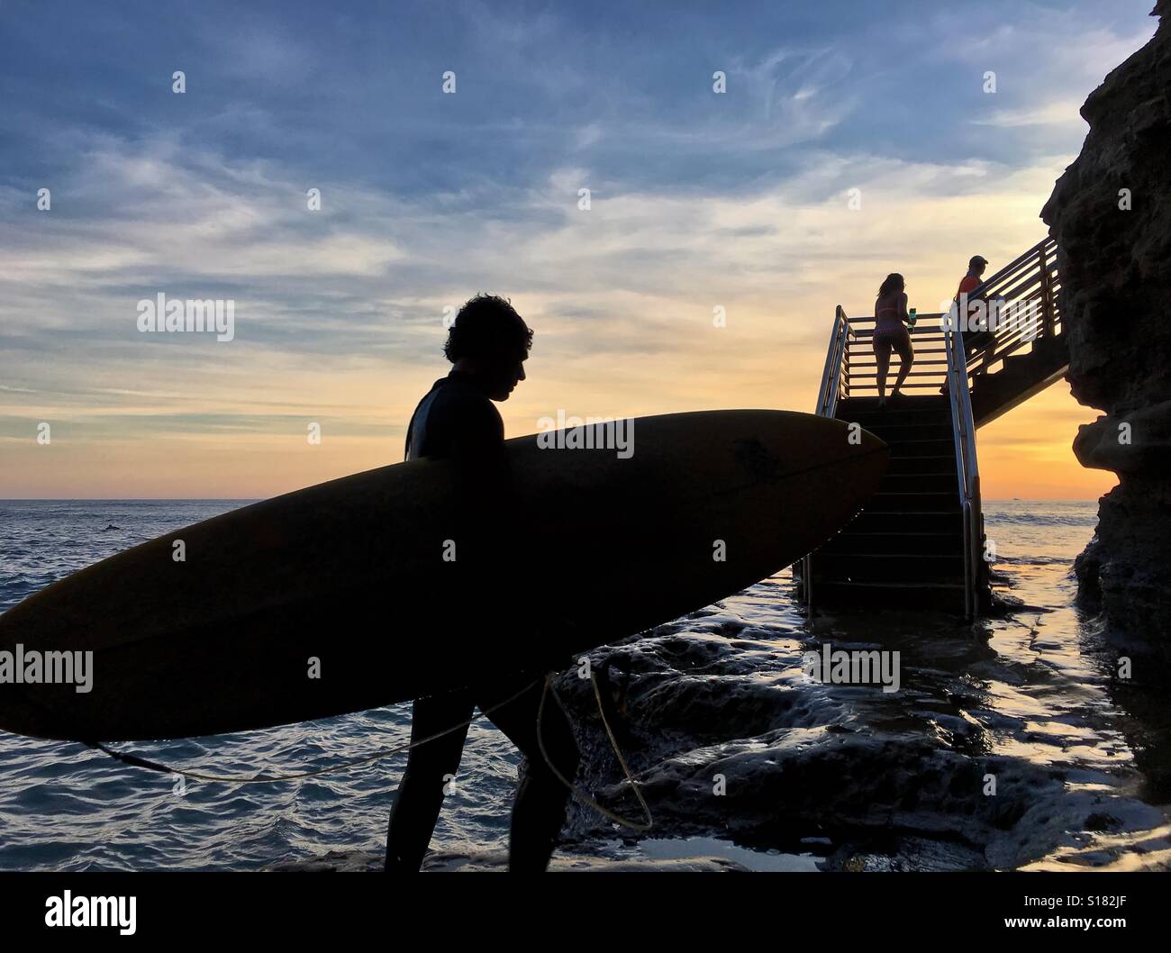 Silhouette of a surfer hold a surfboard along rugged coastline. Dusk at ...