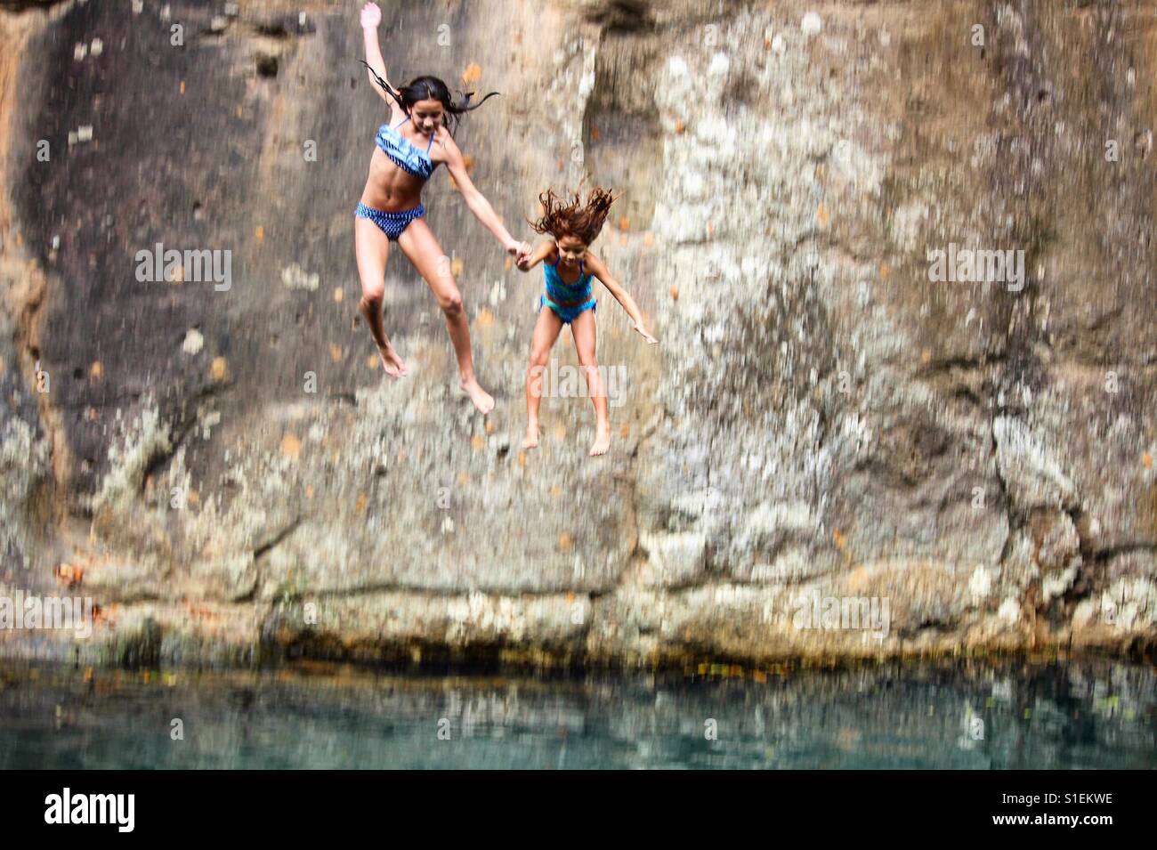 Two Girls Holding Hands While Jumping Off Of A Cliff Into A River