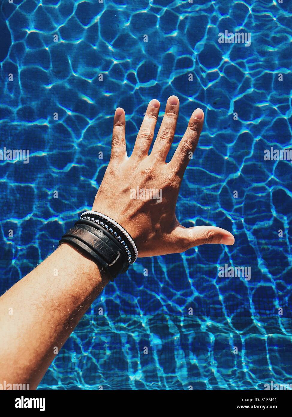 A male hand wearing bracelets outstretched over a blue swimming pool ...