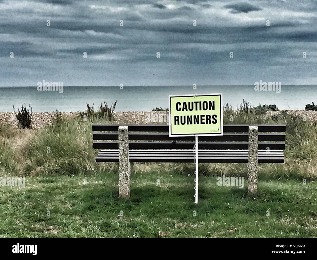 Caution Runners sign leaning against bench on Greensward facing the sea ...