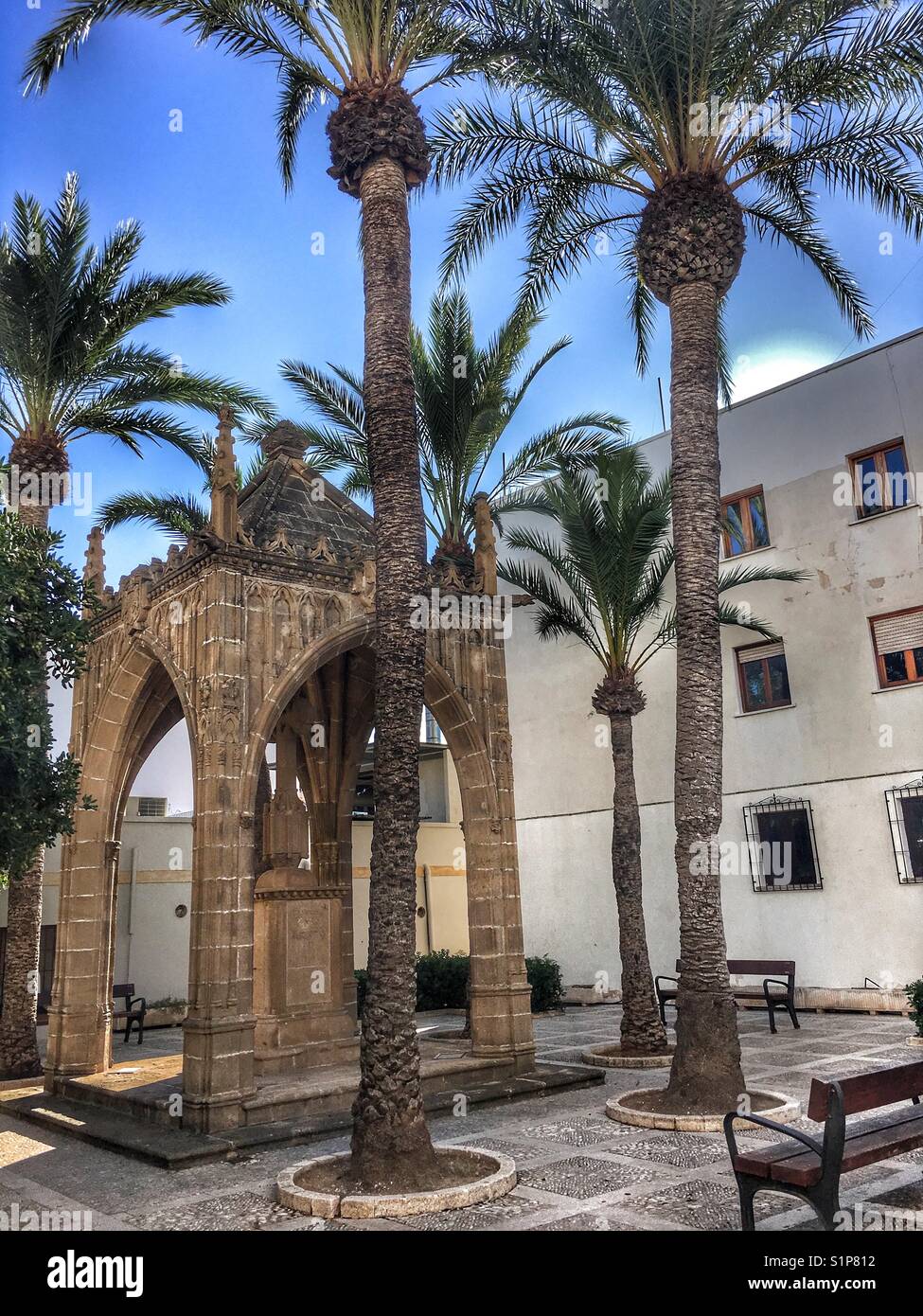 Typical small Spanish square with shady palms and a religious monument ...