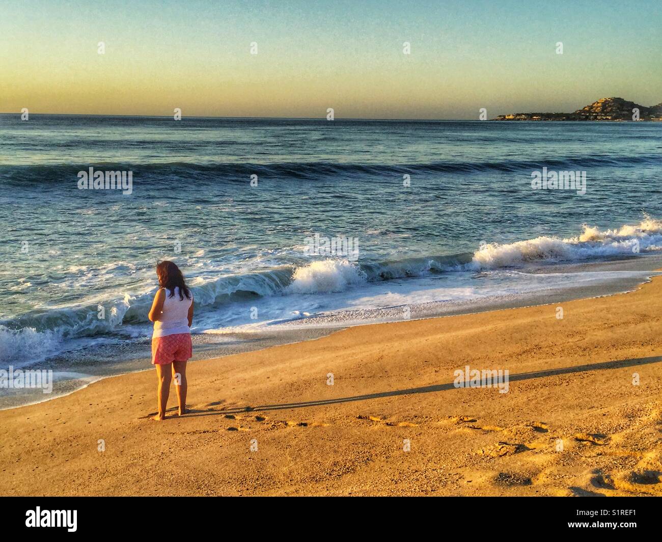 Woman stands alone on beach looking at the sea as the sun sets Stock ...