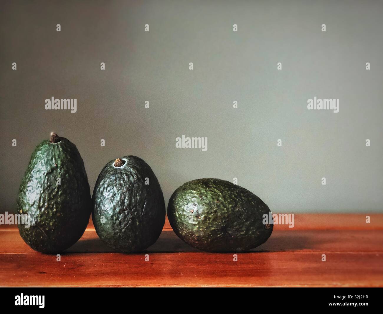 Still life of three avocados on wooden surface with plain grey ...