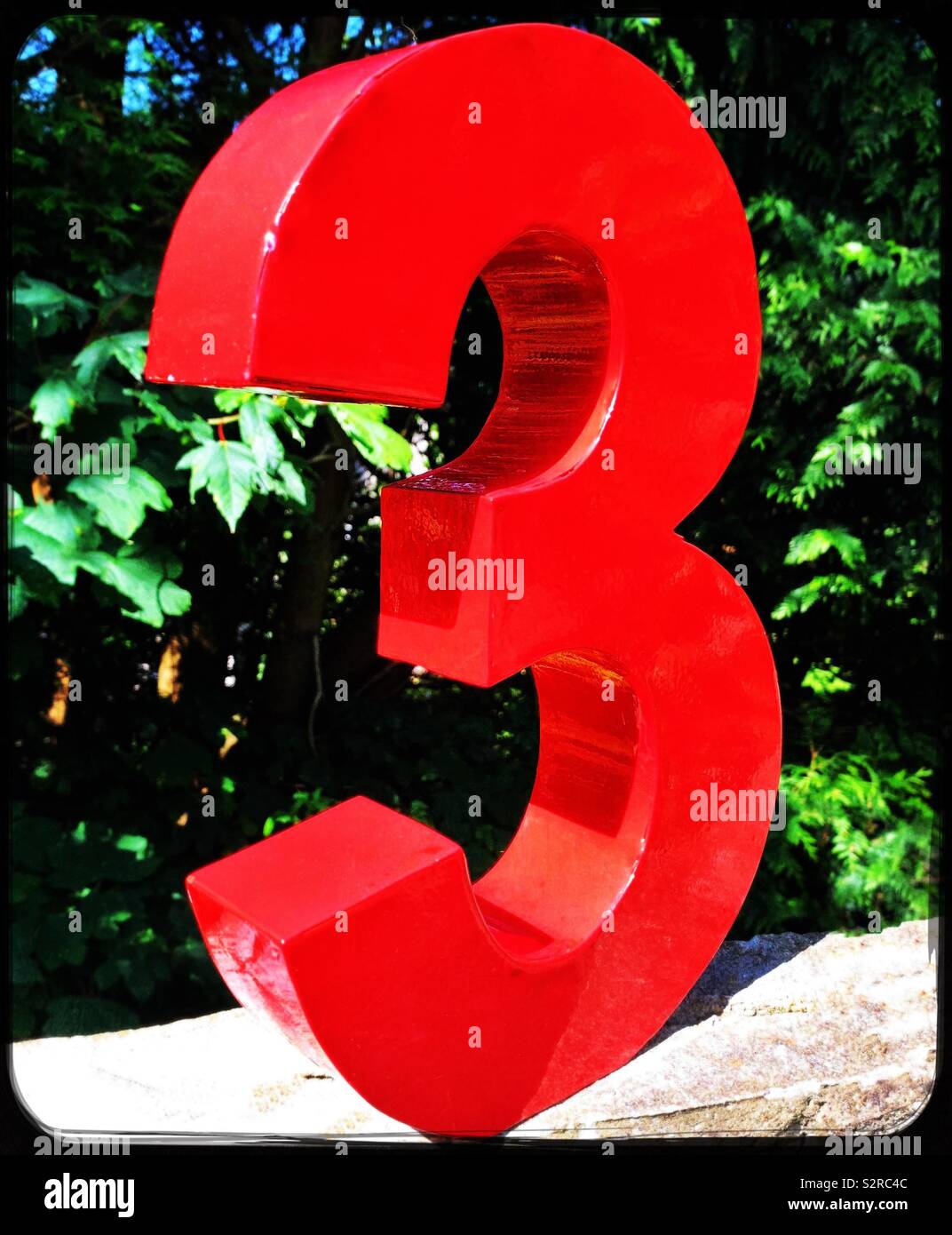 Red box metal number three on wall in garden with trees in background ...