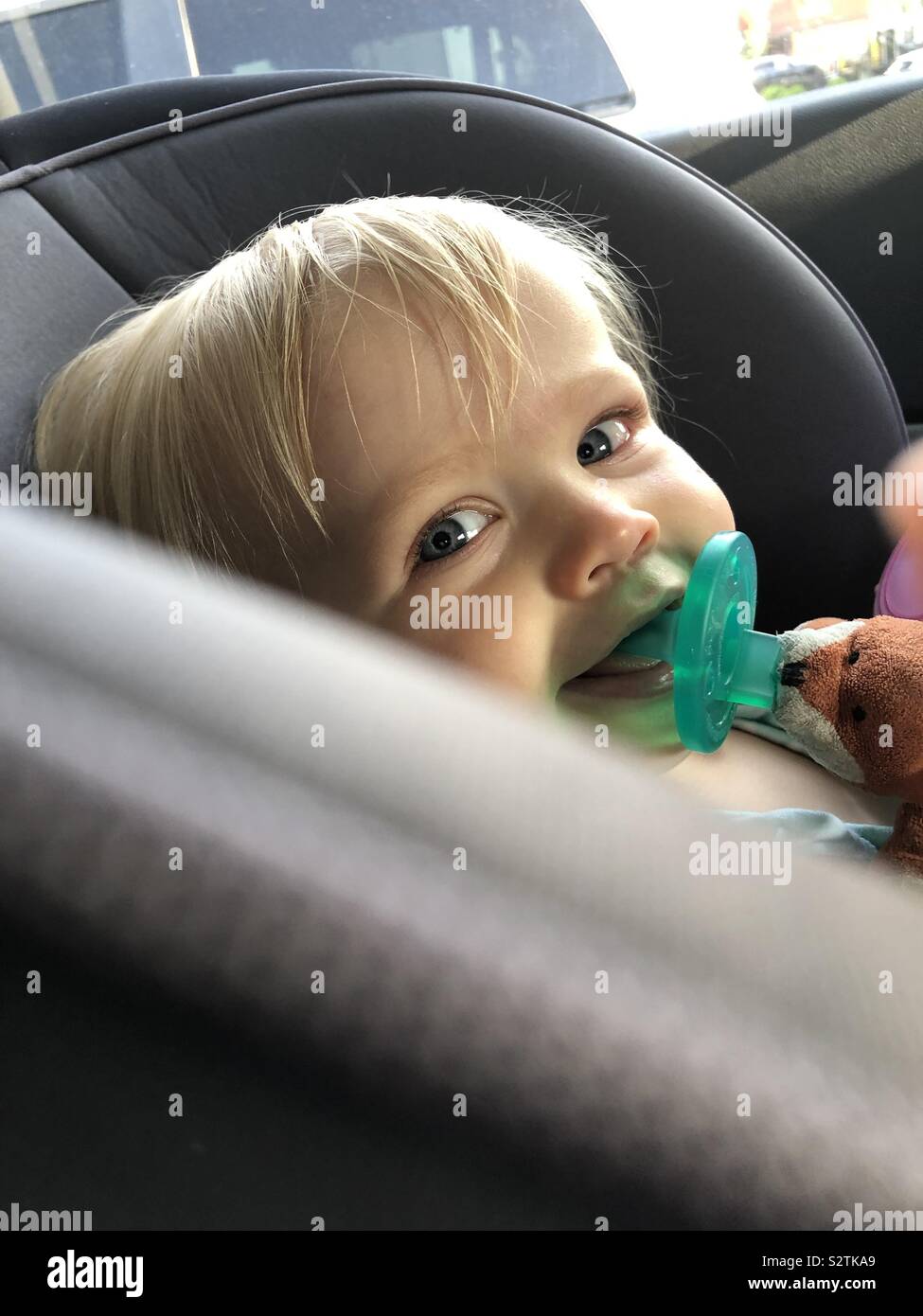 A toddler smiling with a pacifier in her mouth while seated in a car ...