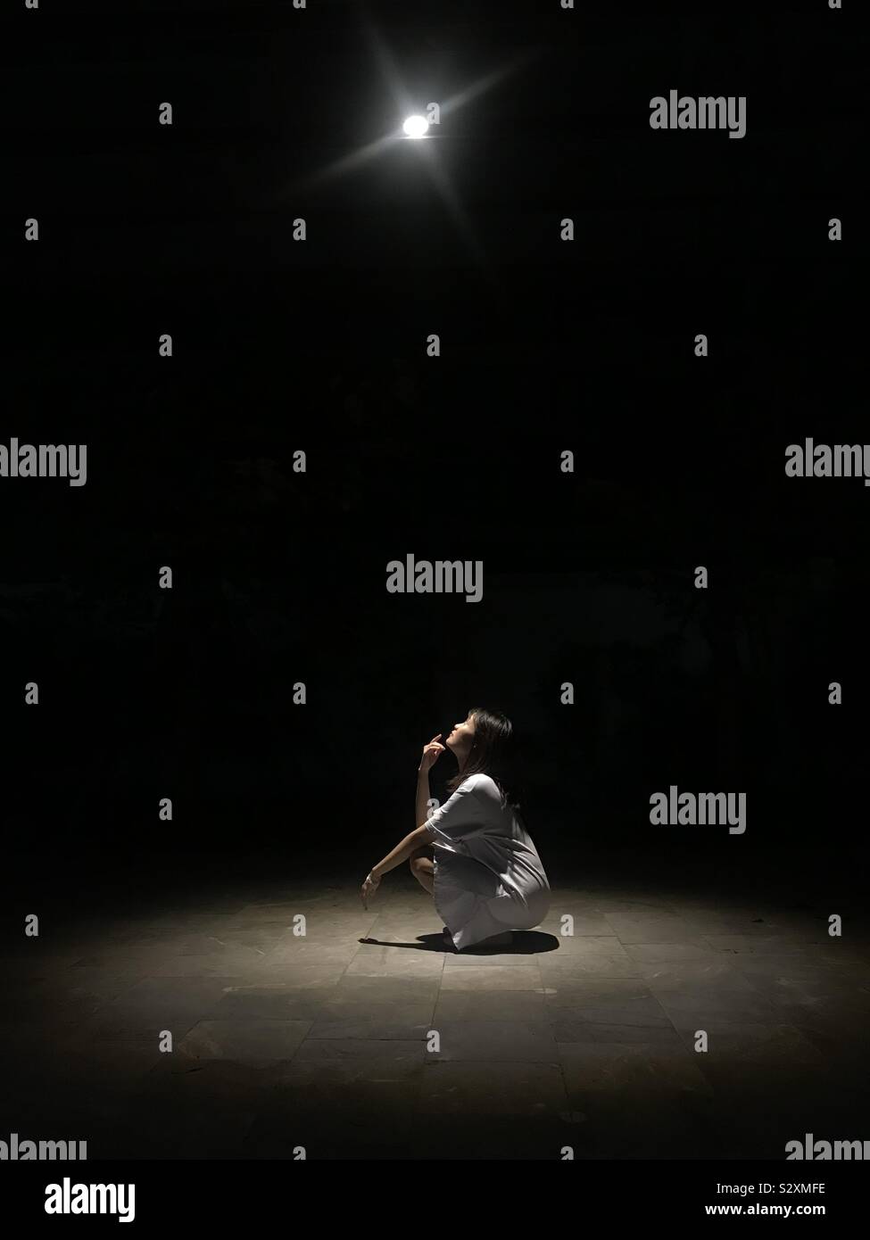 A lonely girl sitting at a spotlight looking up at a loght above Stock ...