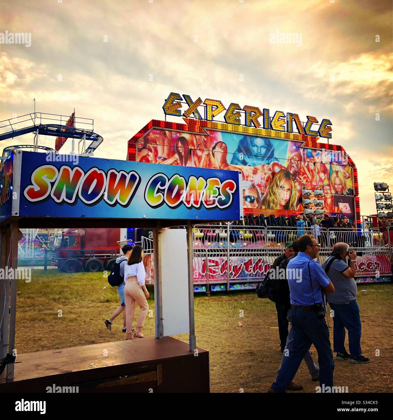 People at a colourful fairground during sunset, with snow cones stall ...