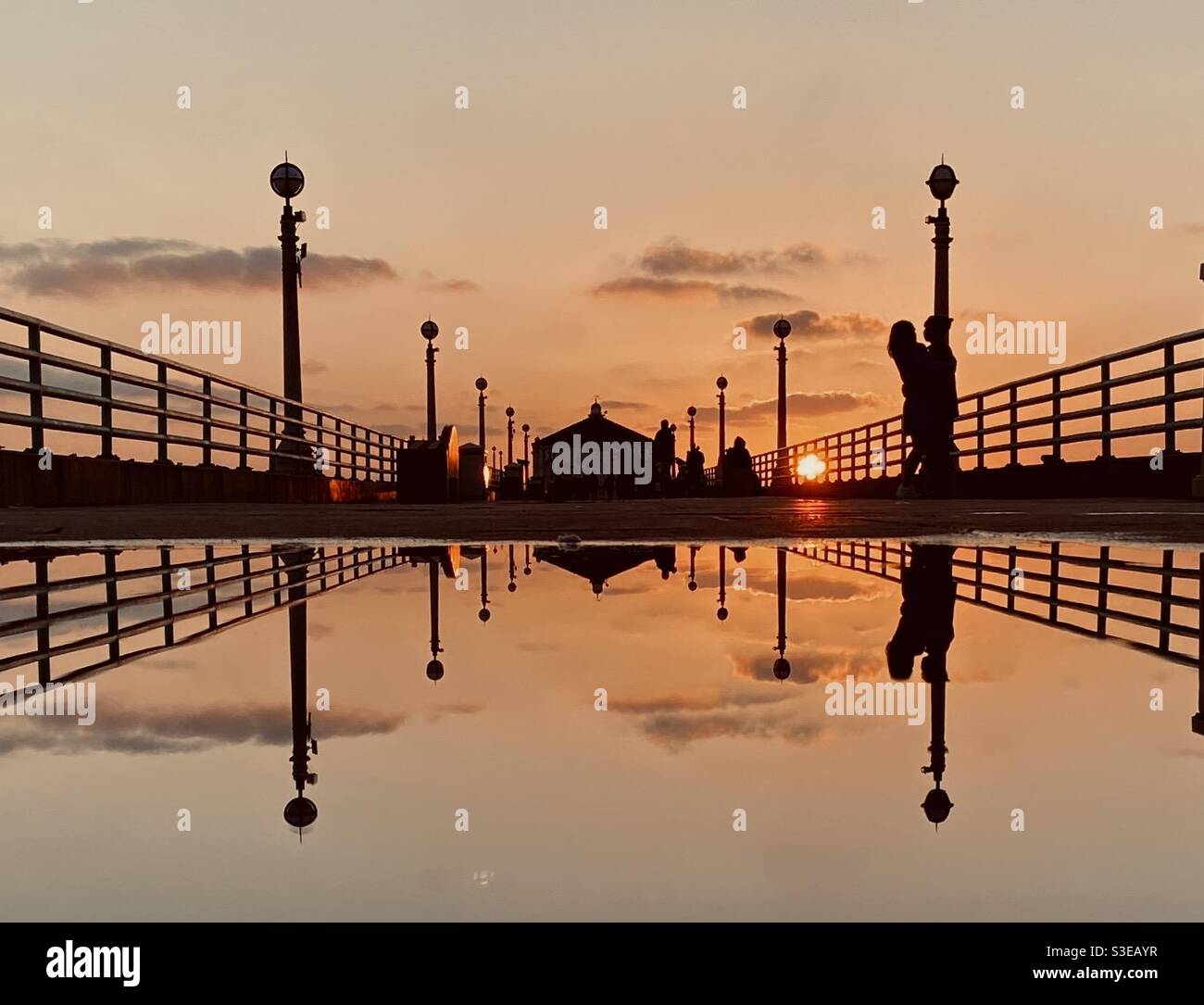 A puddle of water reflexes the Manhattan Beach pier as a couple hugs at ...
