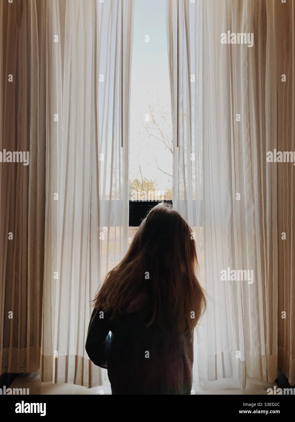 Small child looking out a tall window with white curtains in a hotel ...