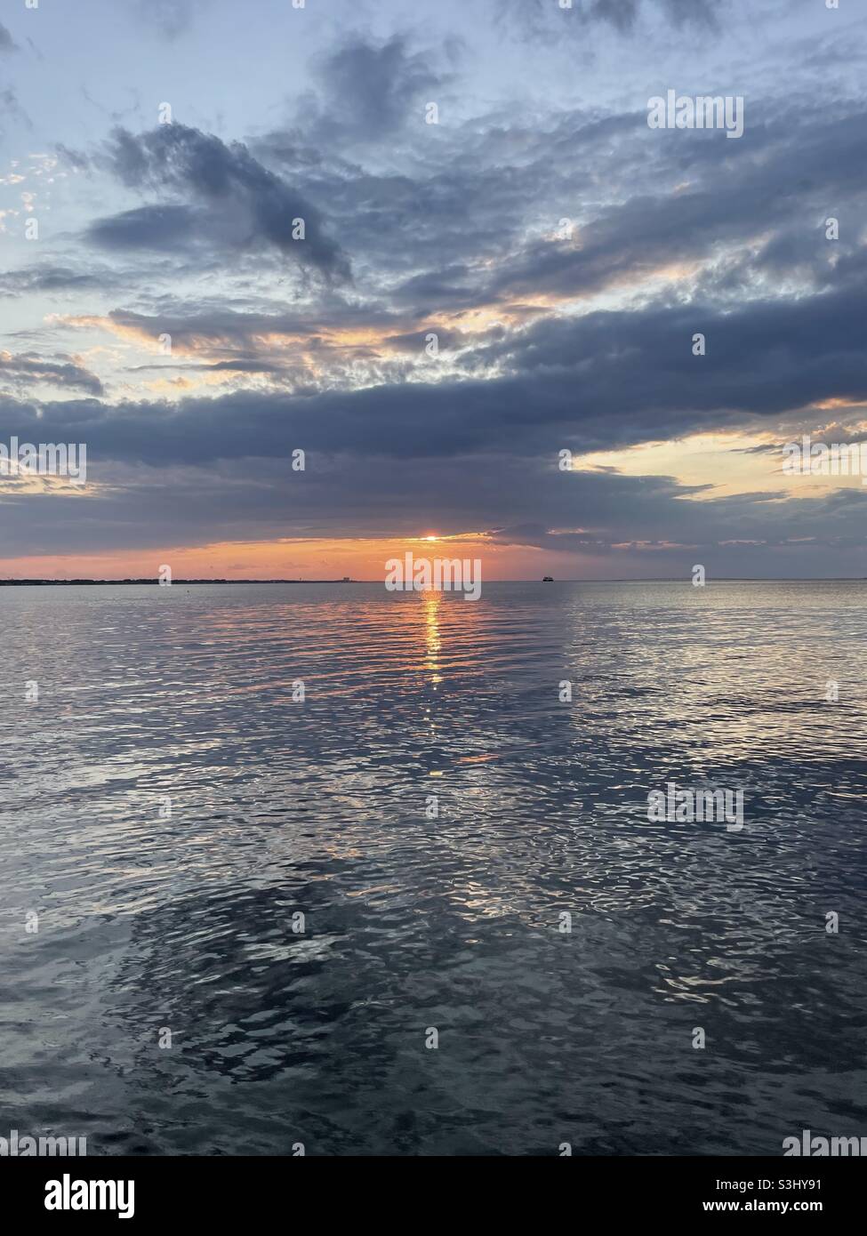 Sun under a large band of clouds at sunset reflecting onto bay water ...